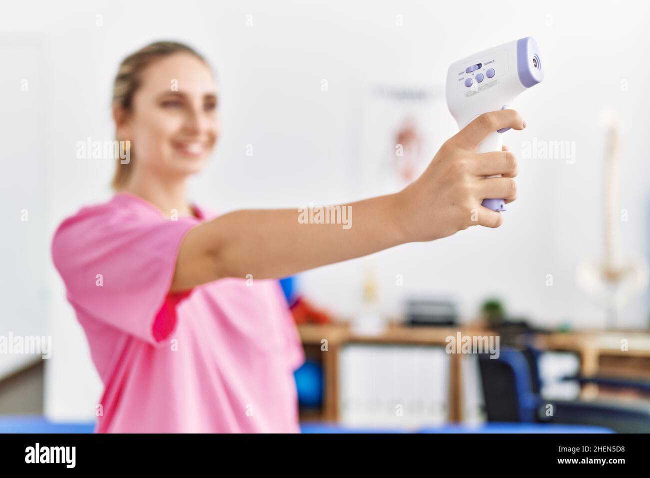 Young blonde woman wearing physiotherapist uniform using thermometer at ...