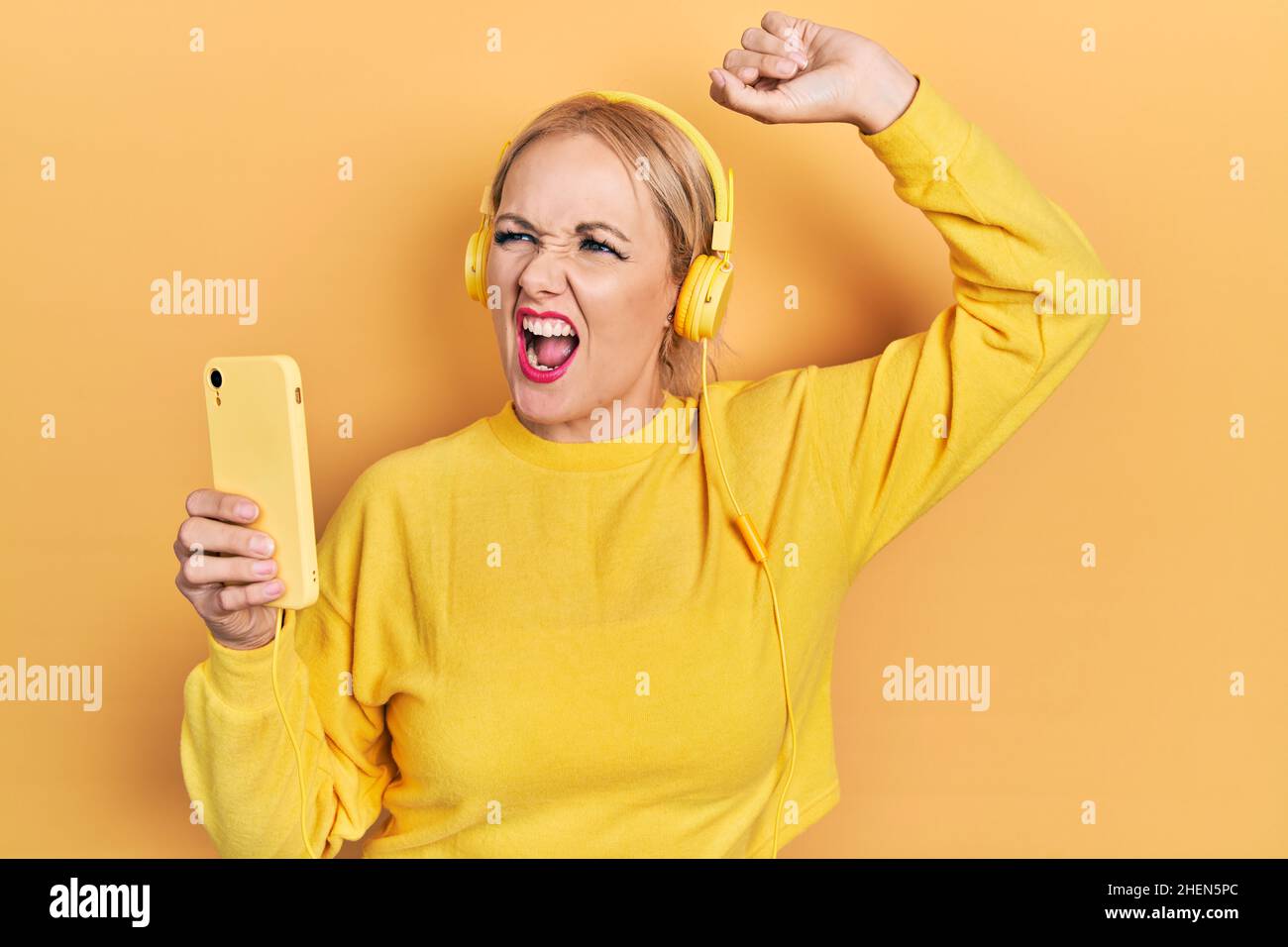Young blonde woman dancing wearing headphones angry and mad screaming ...