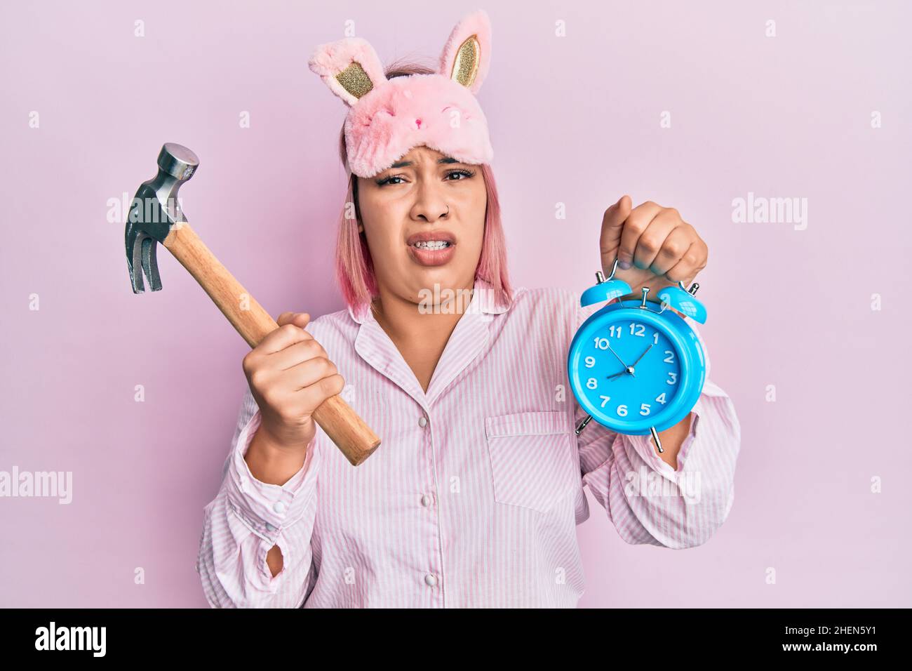 Hispanic woman with pink hair wearing pajama holding hammer and alarm ...