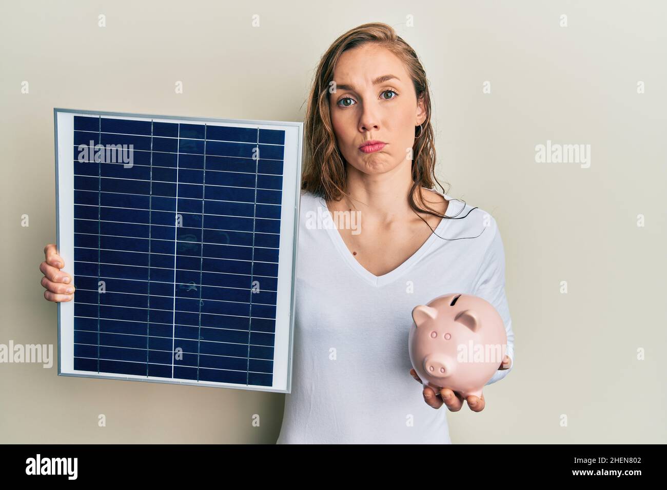 Young blonde woman holding photovoltaic solar panel and piggy bank ...