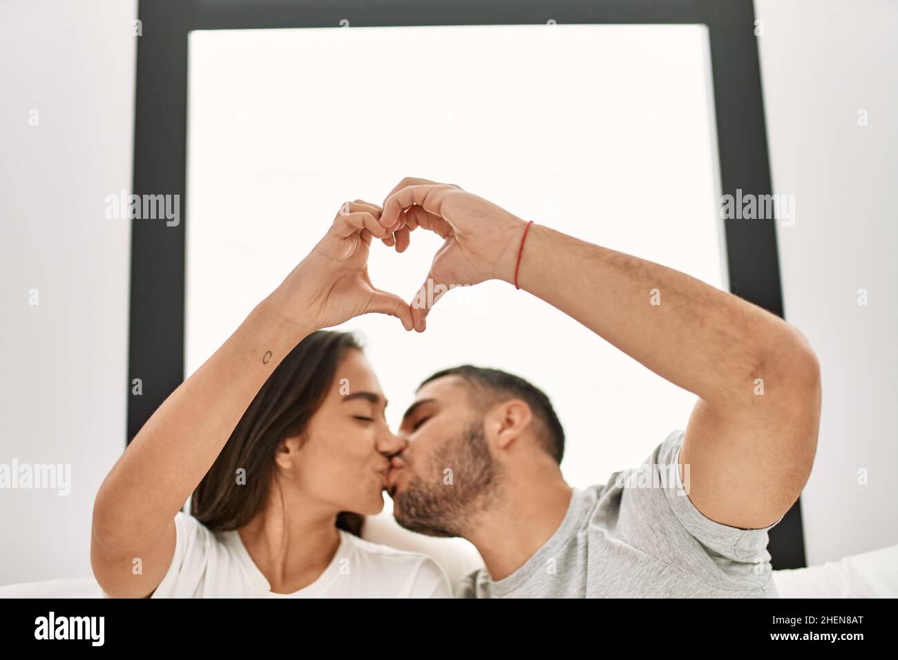 Young hispanic couple doing heart symbol with hands sitting on the bed ...