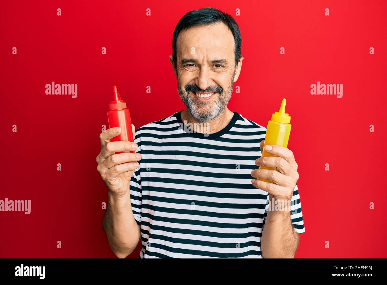 Middle age hispanic man holding ketchup and mustard bottle smiling with ...