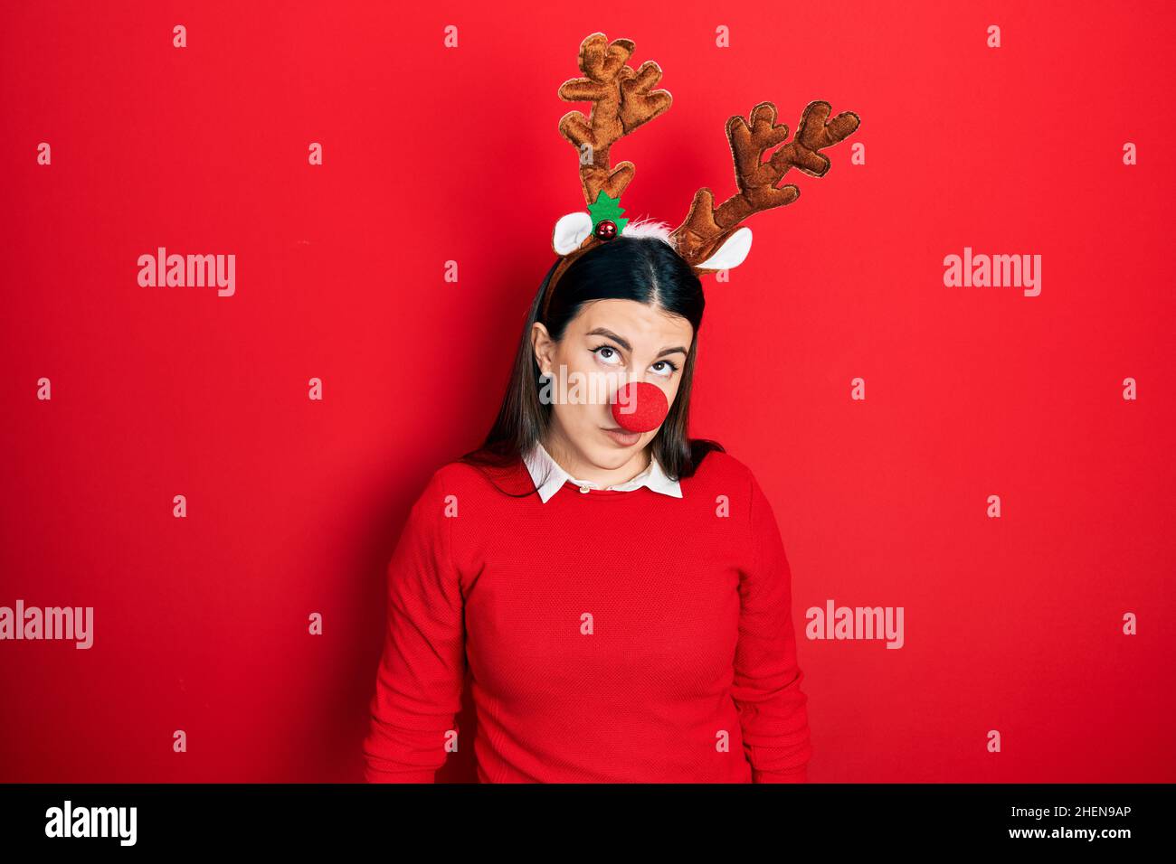 Young hispanic woman wearing deer christmas hat and red nose looking ...