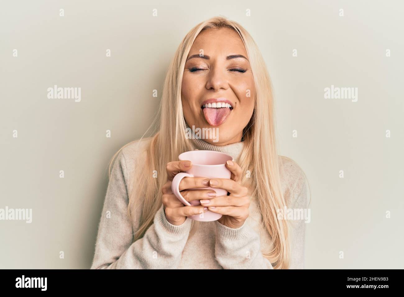 Young blonde woman drinking a cup of coffee sticking tongue out happy ...
