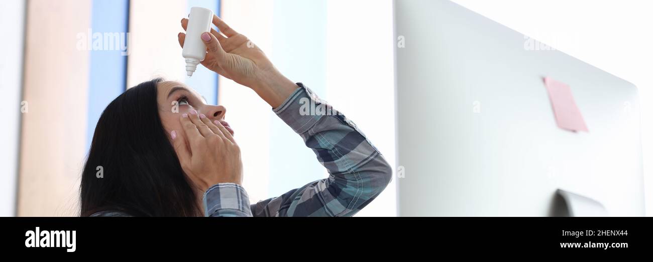 Woman drips eye drops into eyes while sitting at workplace at computer ...