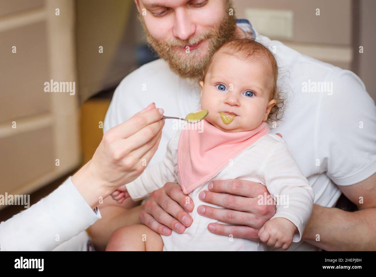 Father and mother feed their daughter fruit puree in the kitchen from a ...