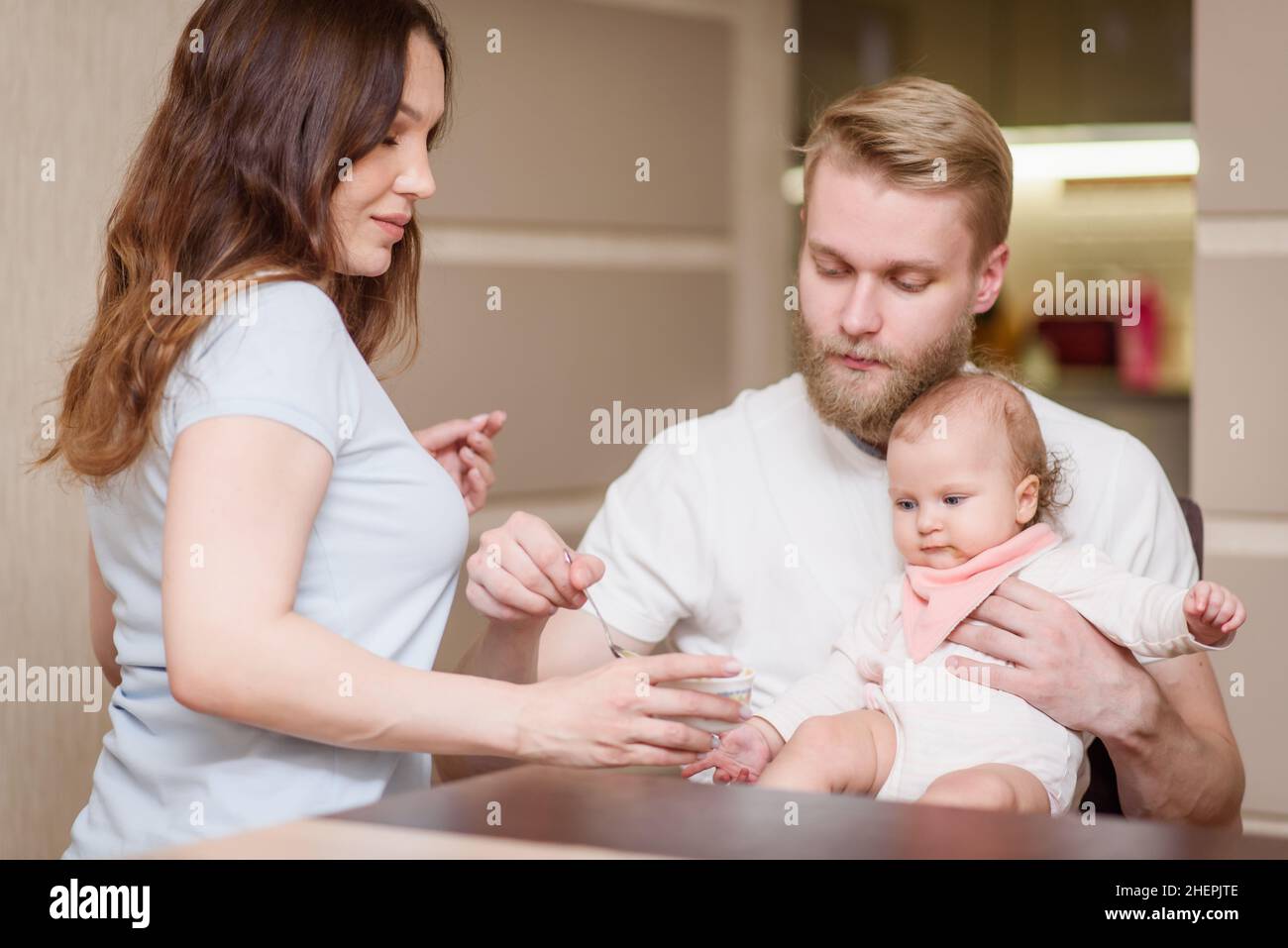 Father and mother feed their daughter fruit puree in the kitchen from a ...