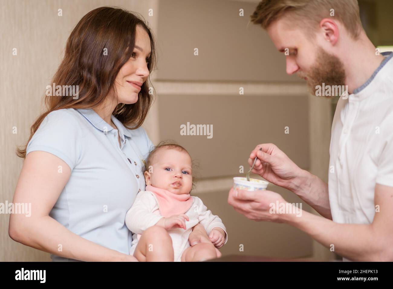 Father and mother feed their daughter fruit puree in the kitchen from a ...