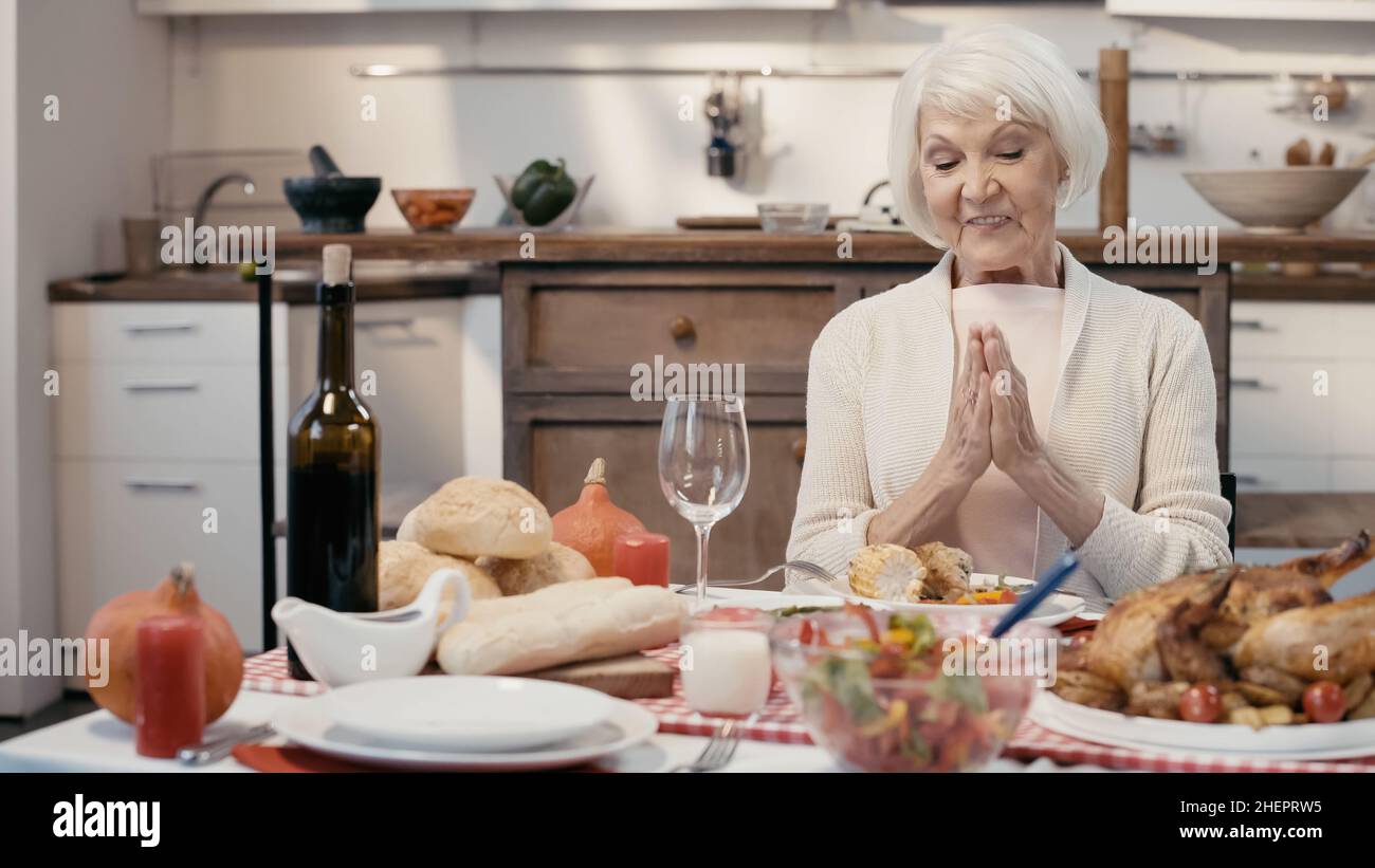 smiling senior woman praying before thanksgiving dinner near roasted ...