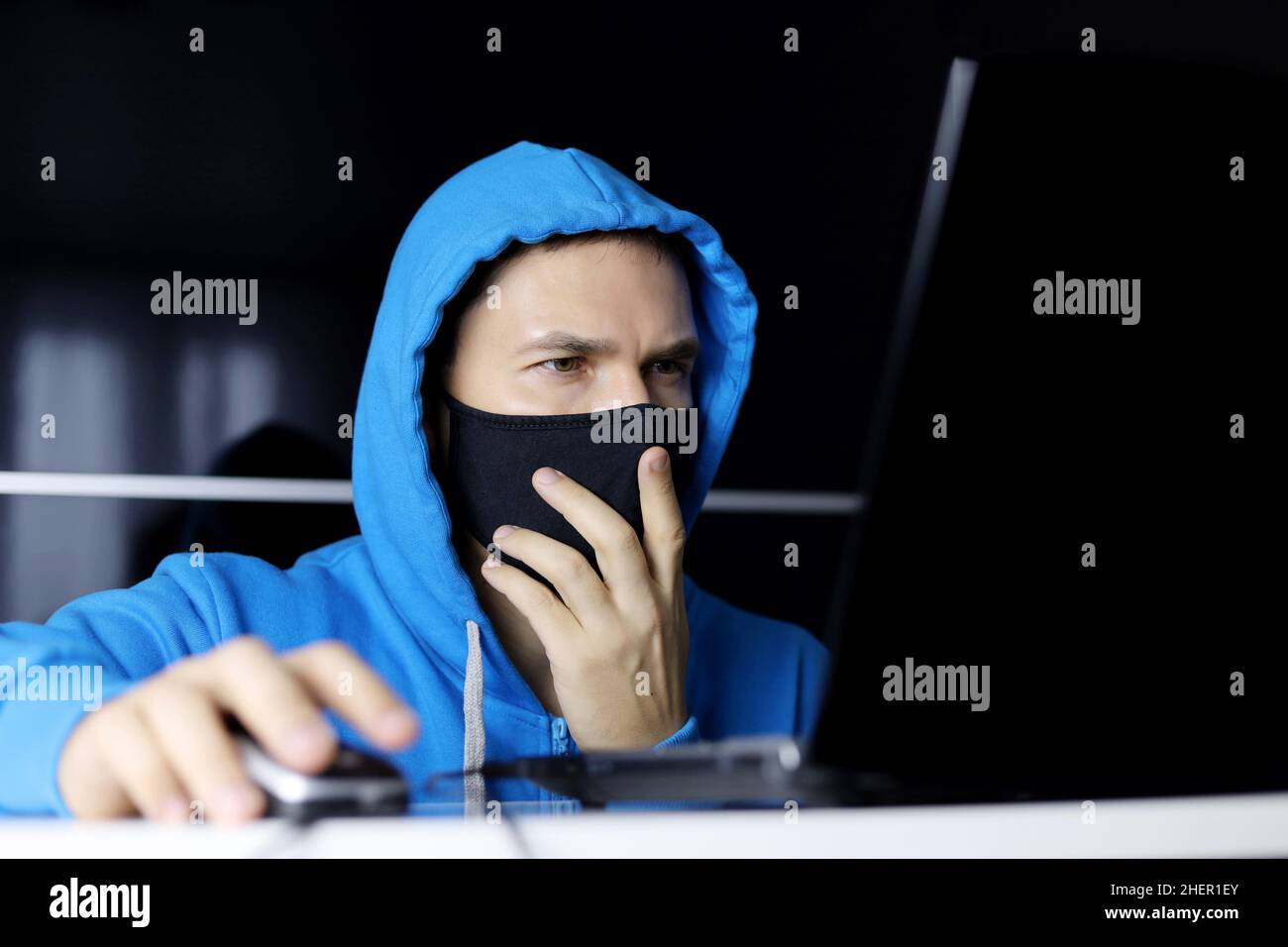 Man in mask and blue hoodie sitting at laptop with a thoughtful face ...