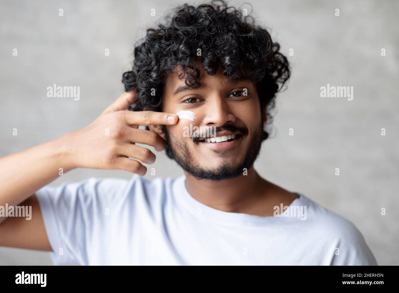 Handsome indian man applying moisturising cream on face while standing ...