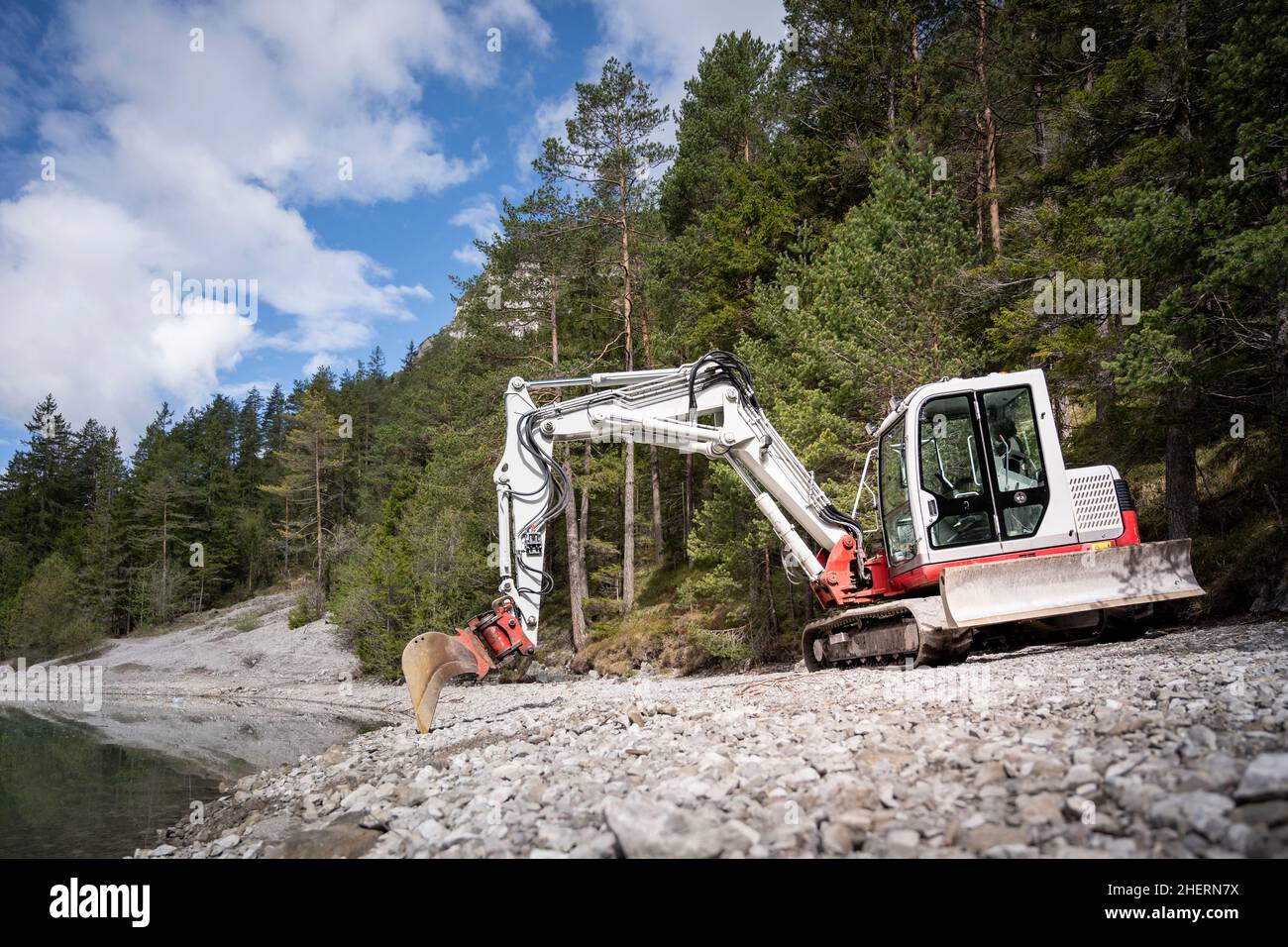 small shovel digger standing on gravel next to lake and forest with ...