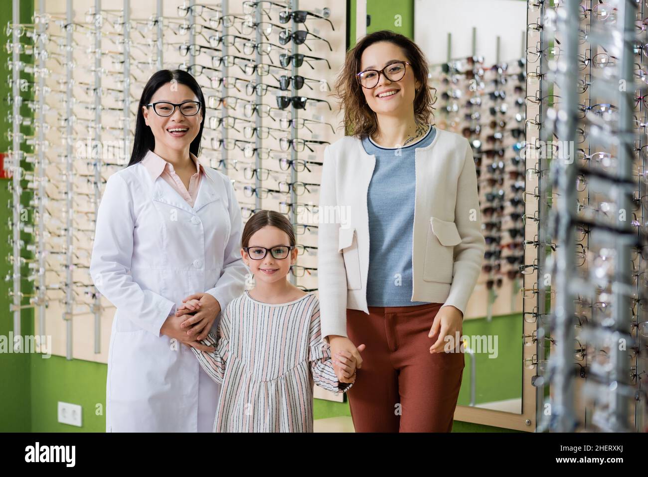 happy girl in eyeglasses holding hands with mom and asian oculist while ...