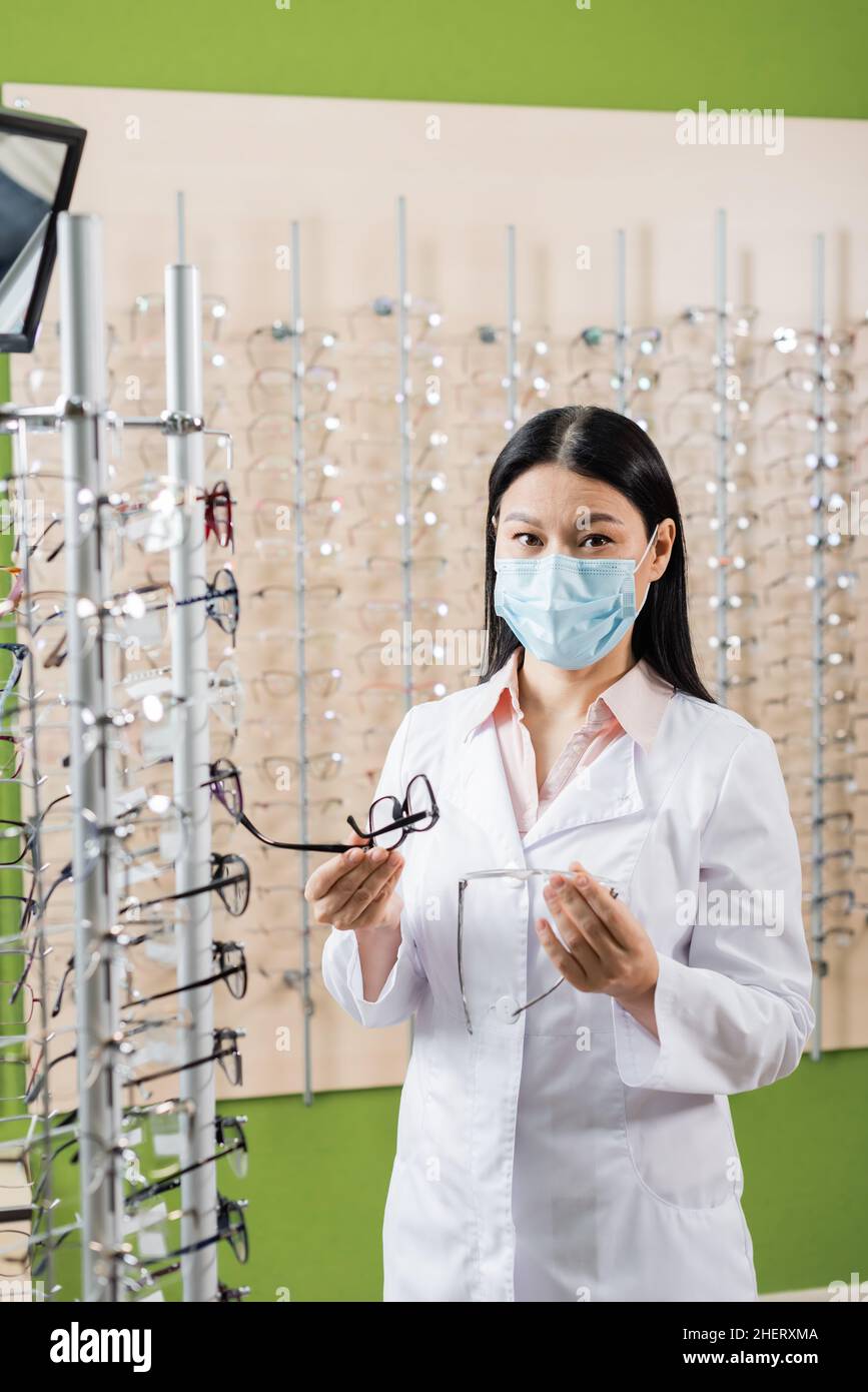 asian oculist in medical mask comparing eyeglasses while working in ...