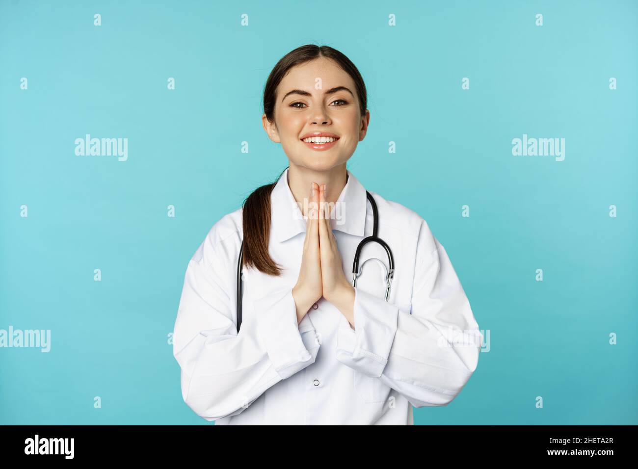 Smiling hopeful young woman doctor, holding hands in begging, thank you ...