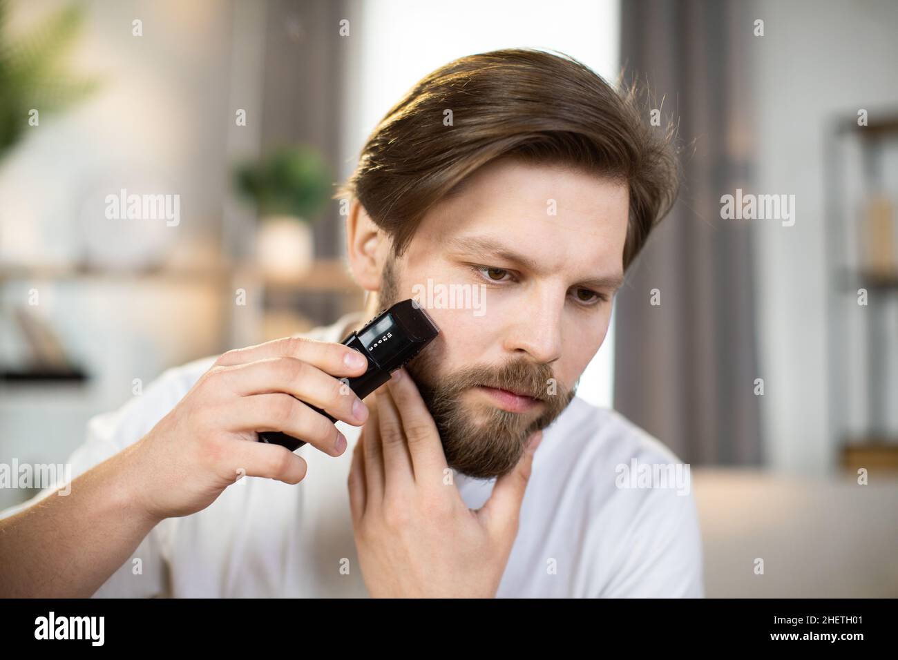 Positive attractive young guy with beard trimming his beard on his cheek with an electronic ...