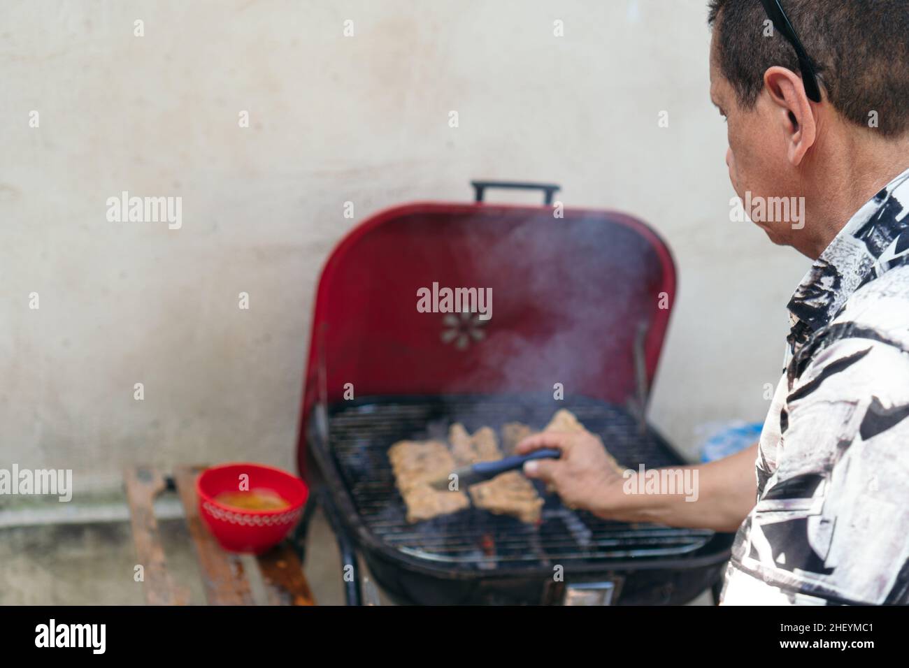 Older man preparing meat on a barbecue grill for his family's lunch in ...