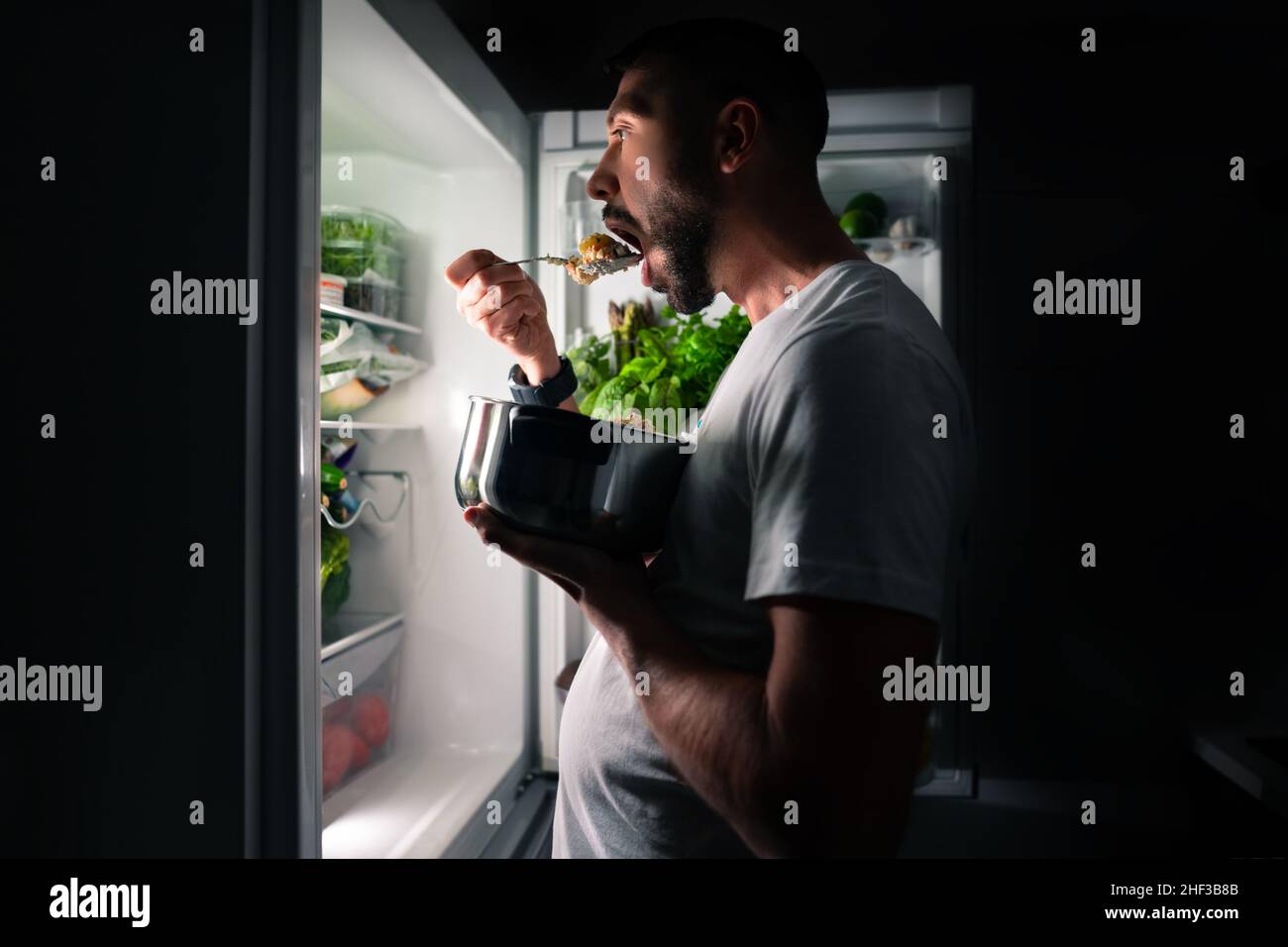 Young hungry man eating food at night from open fridge. Midnight snack ...
