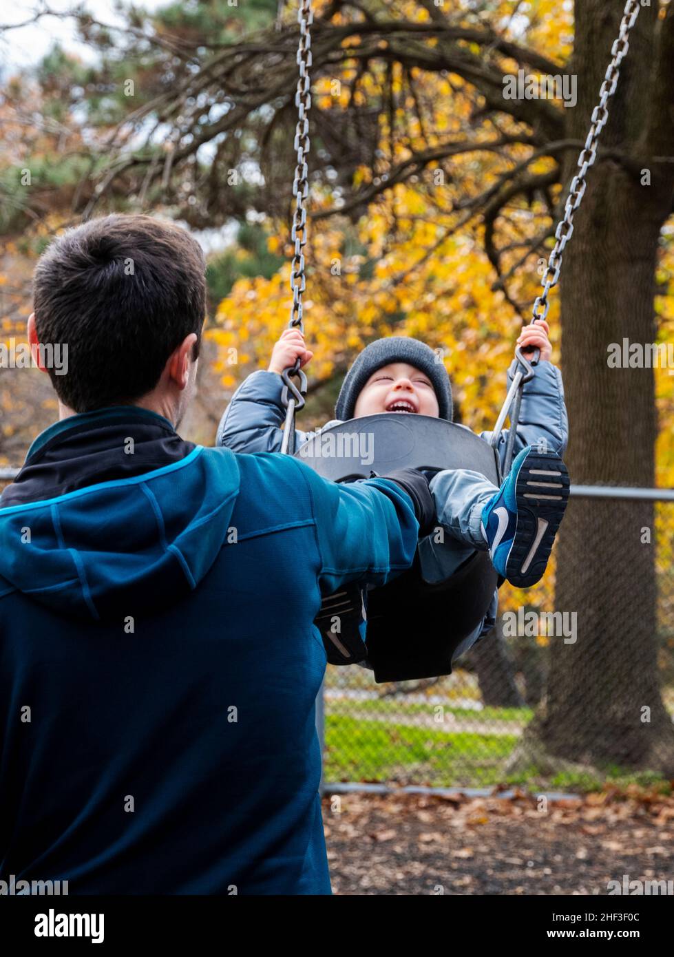 Father pushing two year old boy swinging on city park swing set ...