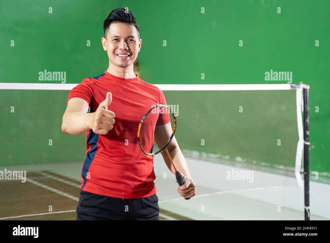 Smiling male badminton player holding racket with thumbs up gesture ...