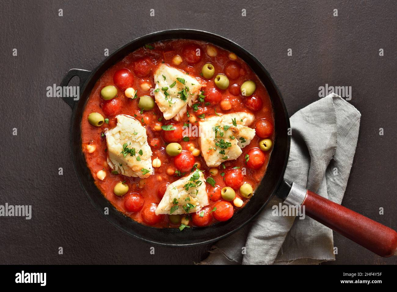 Cod stew with chickpeas, cherry tomatoes and olives in pan over dark ...