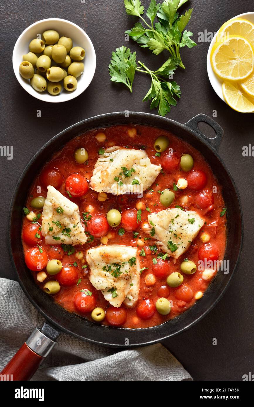 Cod stew with chickpeas, cherry tomatoes and olives in pan over dark ...