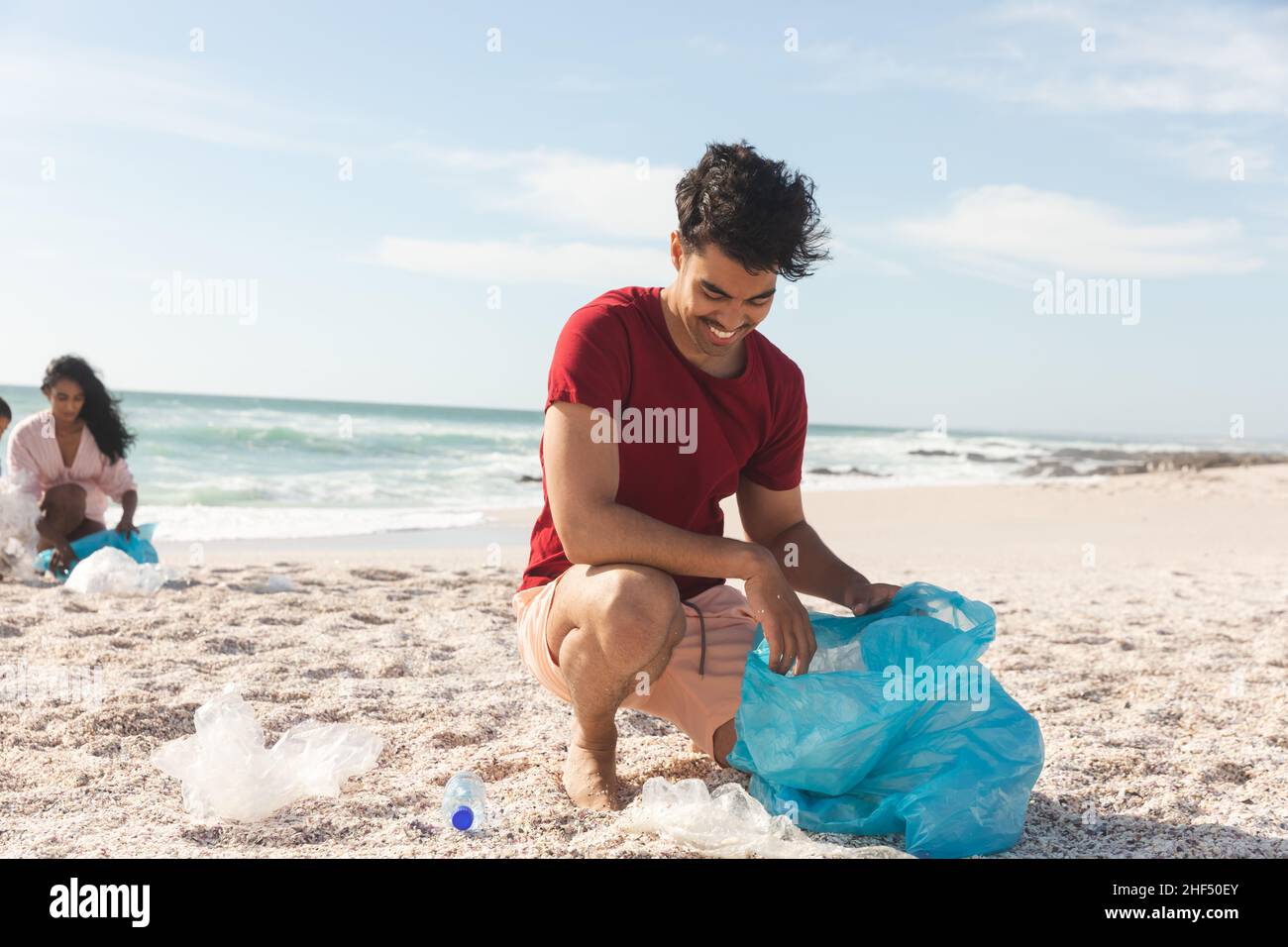 Smiling biracial man collecting garbage in blue plastic bag from sand ...