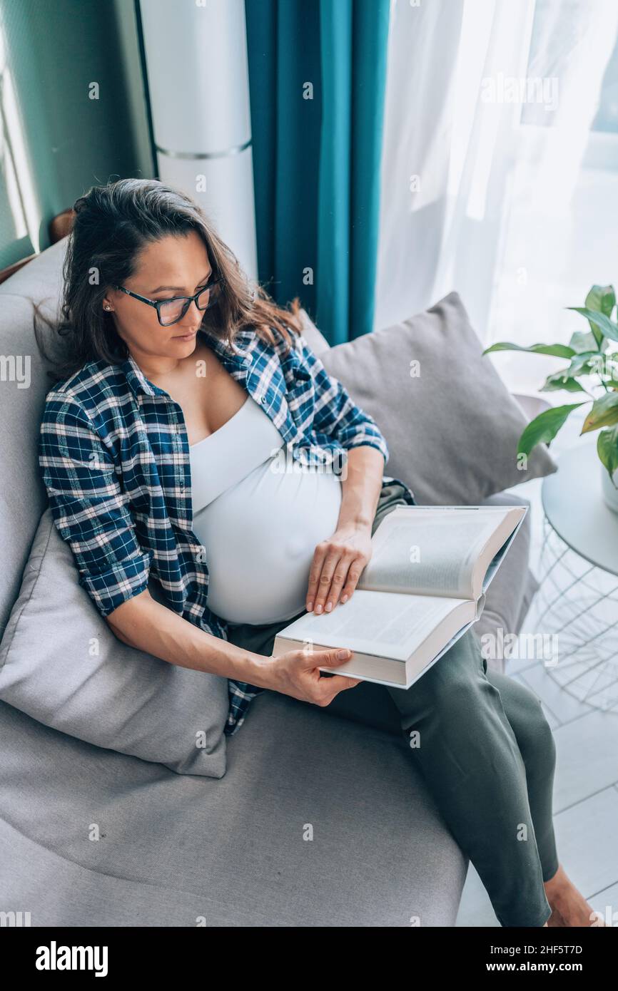 Middle Age Brunette Pregnant Woman In Glasses Read Book Sitting On Grey