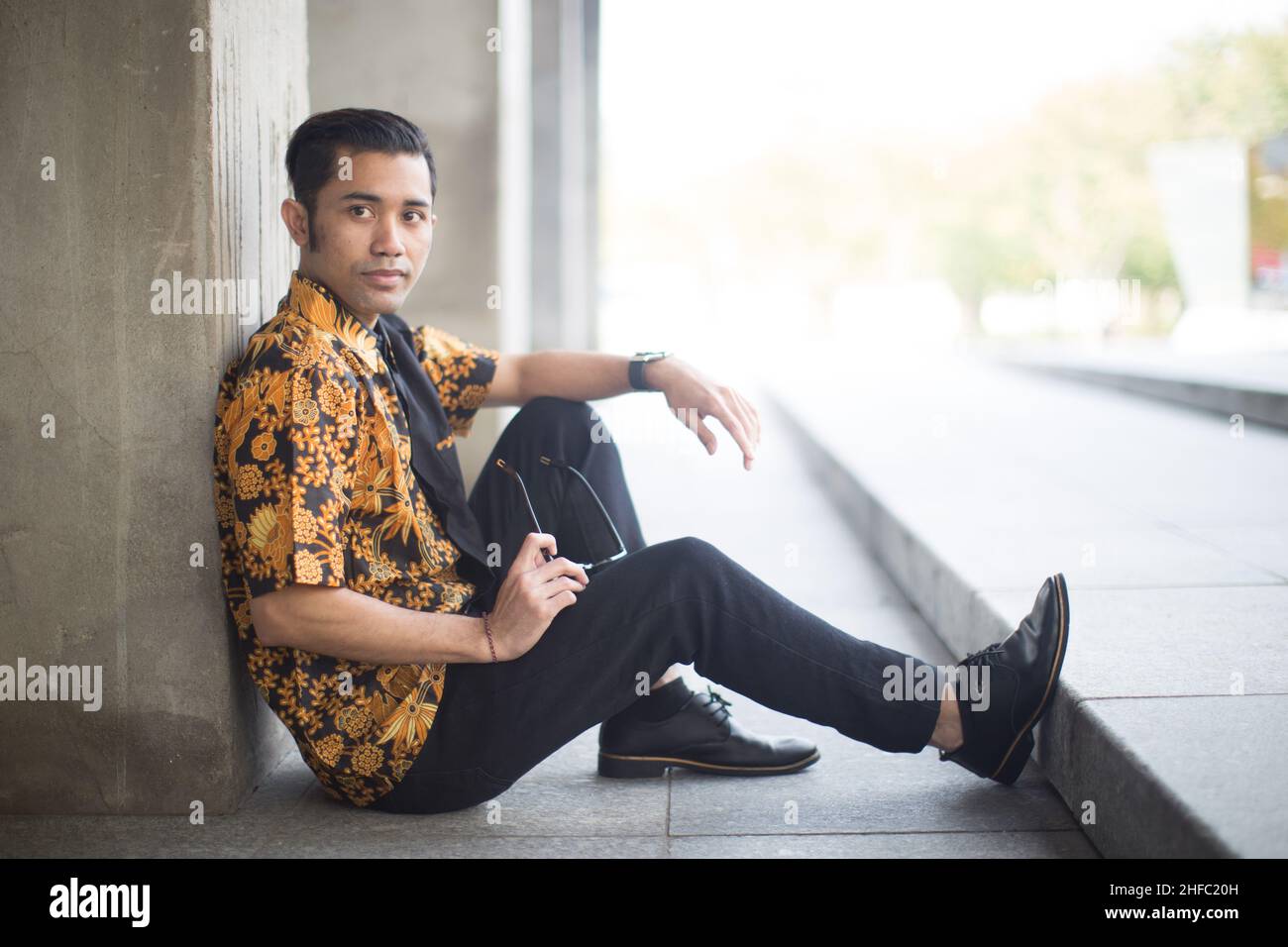 A male model dressed in Indonesian Batik sits and holds sunglasses ...
