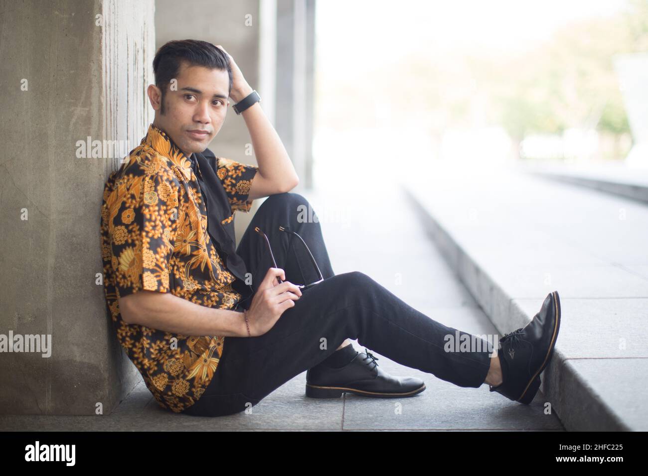 A male model dressed in Indonesian Batik sits and holds sunglasses ...