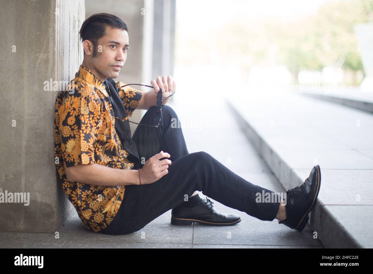 A male model dressed in Indonesian Batik sits and holds sunglasses ...