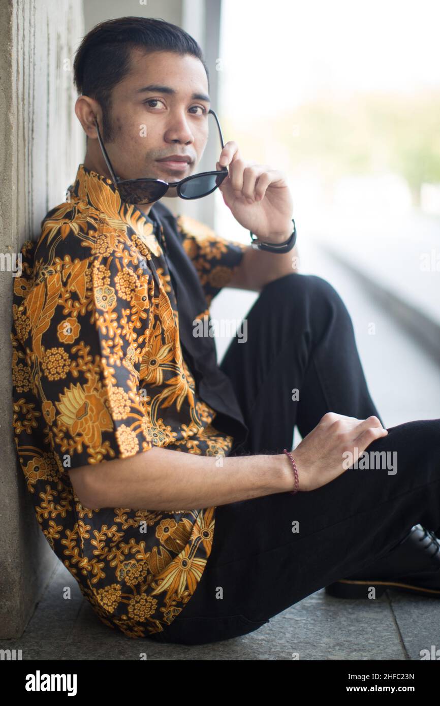 A male model dressed in Indonesian Batik sits and holds sunglasses ...