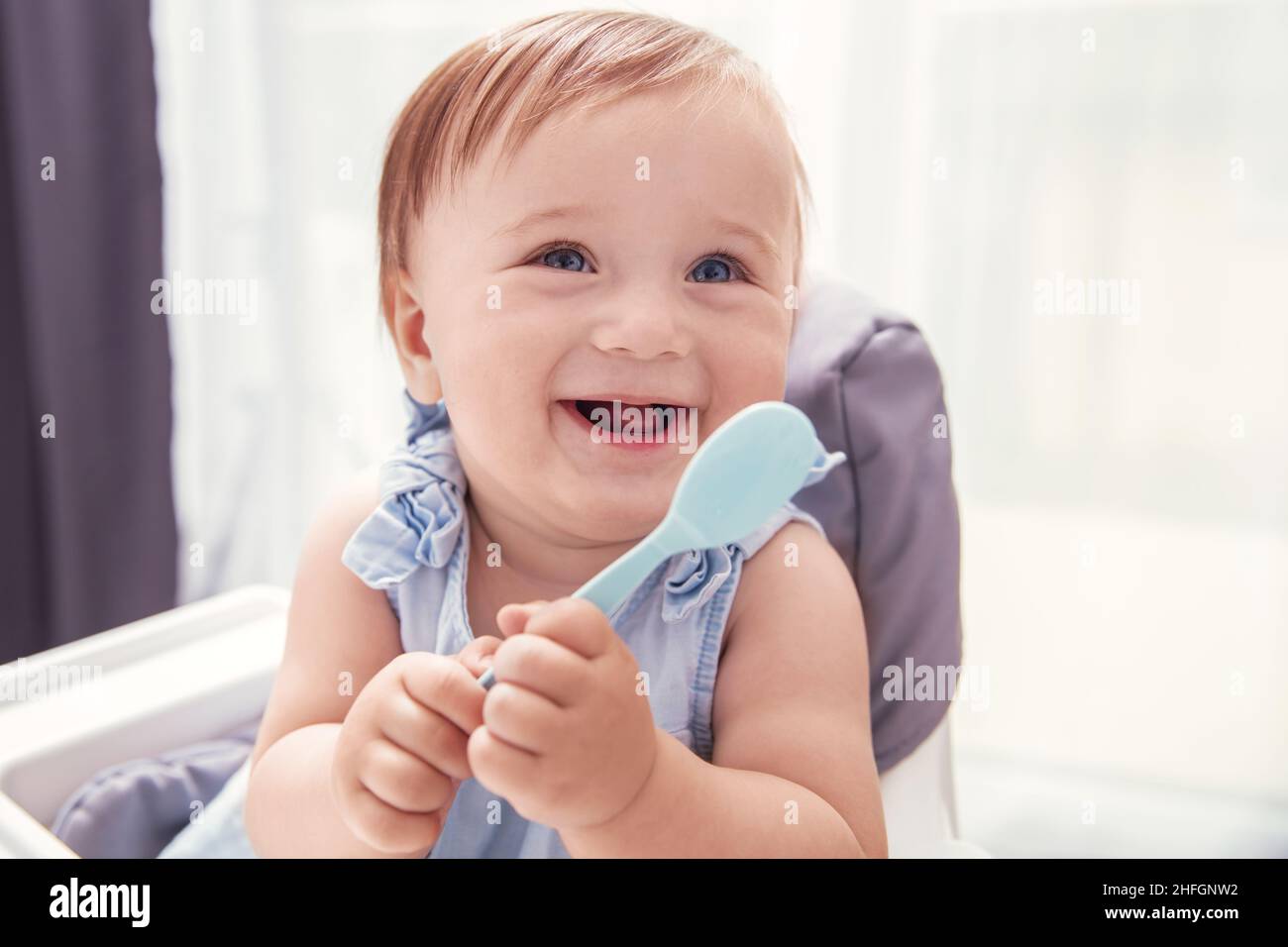 Infant baby girl in blue dress holds baby spoon and smiles Stock Photo ...