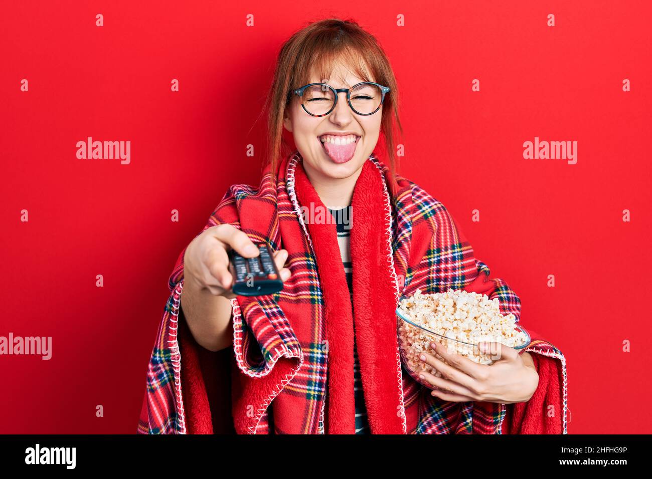 Redhead young woman eating popcorn using tv control sticking tongue out ...