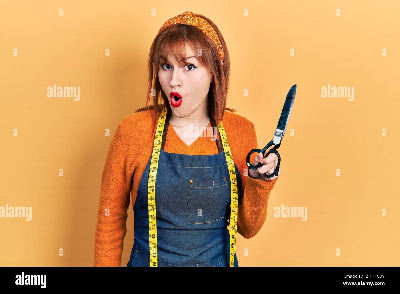Redhead young woman dressmaker designer wearing atelier apron holding scissors scared and amazed ...