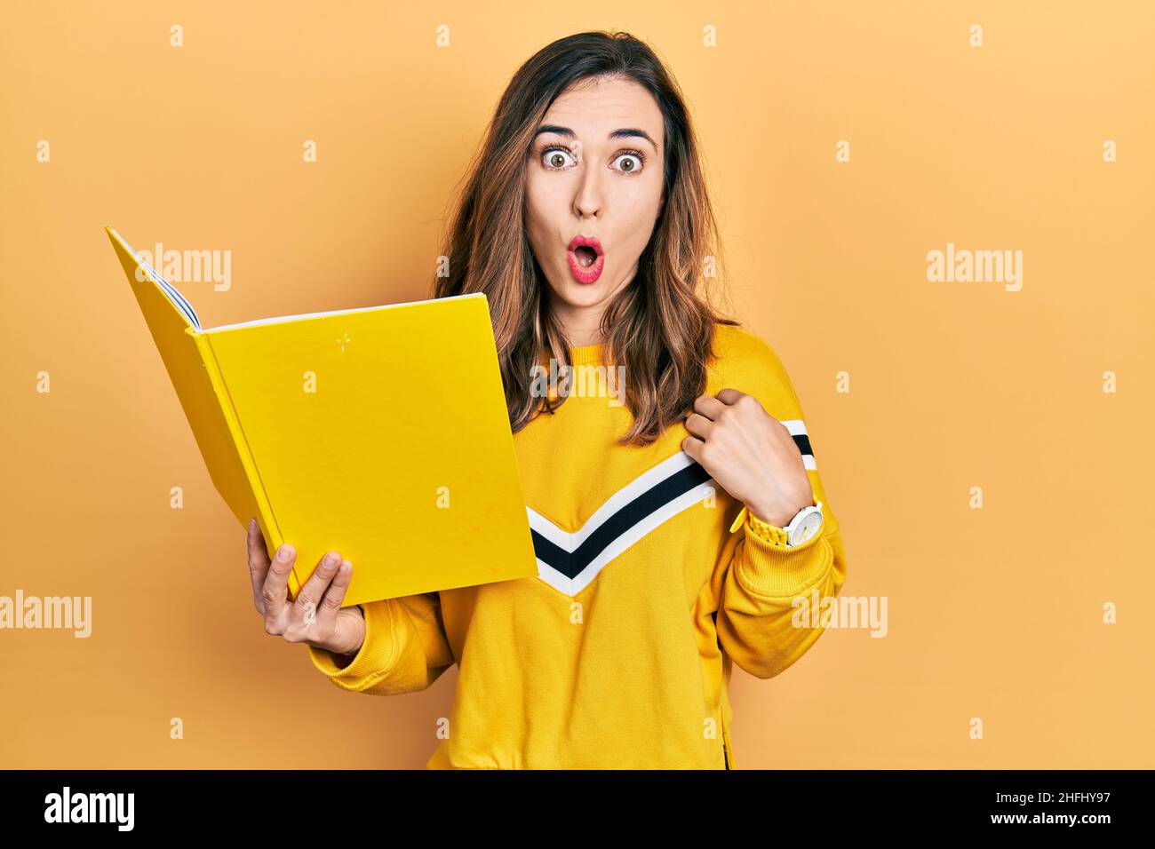 Young hispanic girl reading a book scared and amazed with open mouth ...