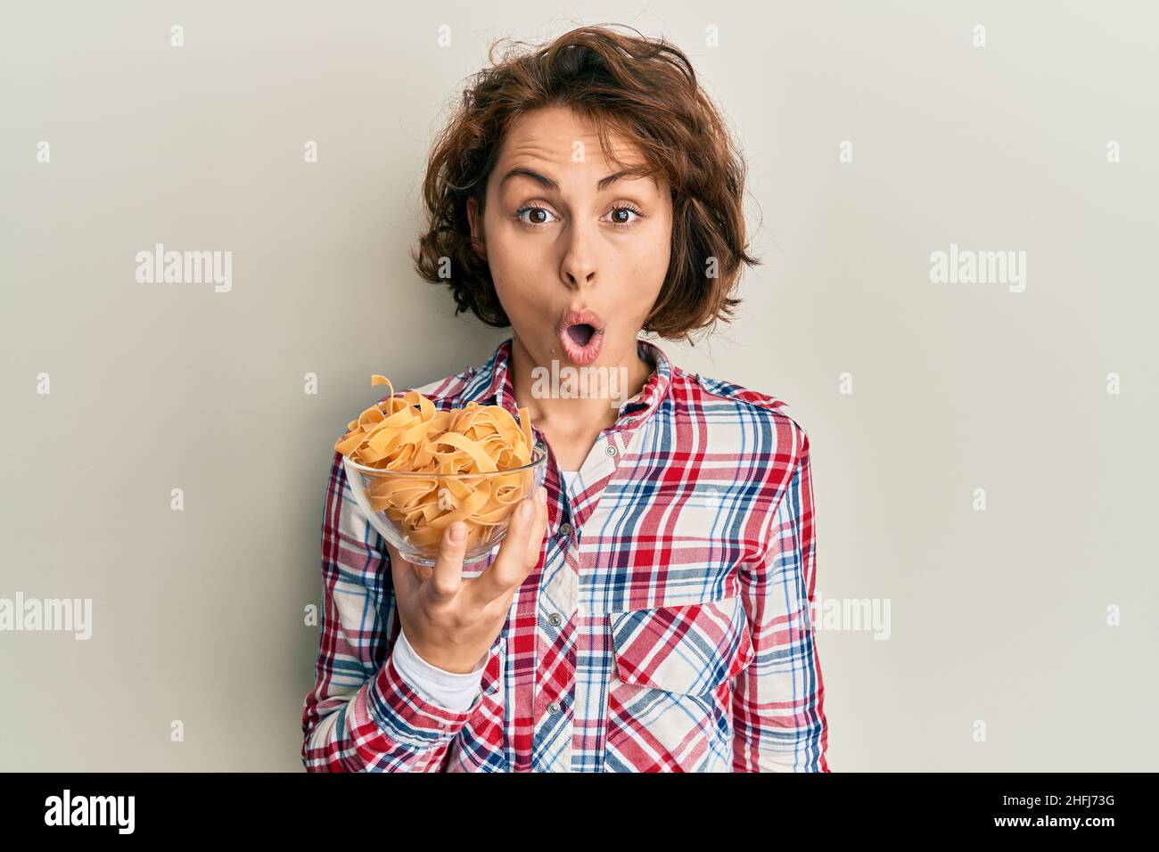 Young brunette woman holding bowl with italian pasta scared and amazed ...