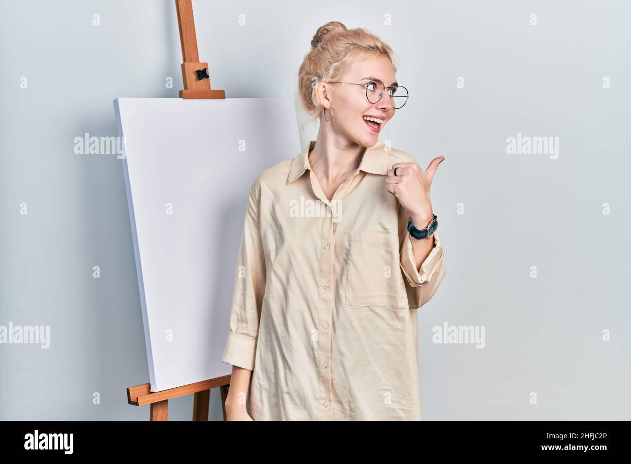 Beautiful caucasian woman with blond hair standing by painter easel ...