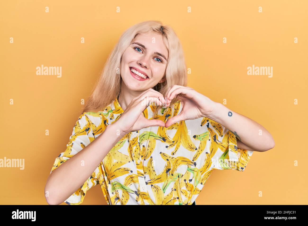 Beautiful caucasian woman with blond hair wearing colorful shirt smiling in love showing heart ...