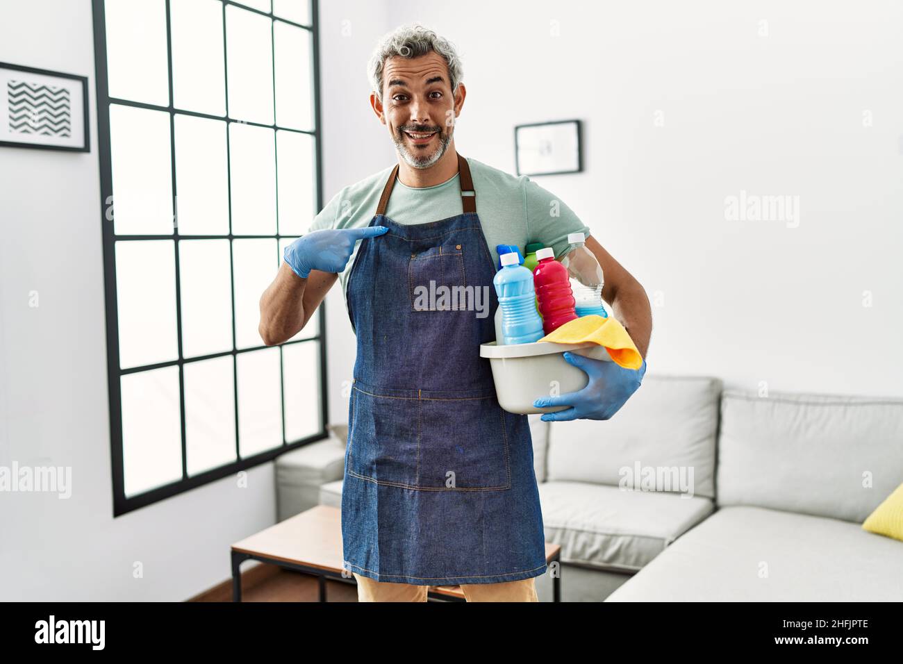 Middle age hispanic man wearing cleaner apron holding cleaning products ...