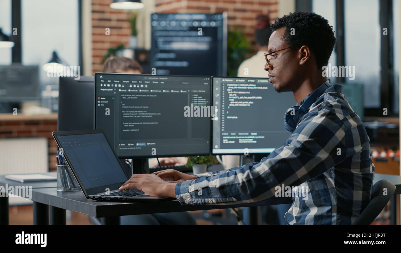 Portrait Of African American Developer Using Laptop To Write Code Sitting At Desk With Multiple