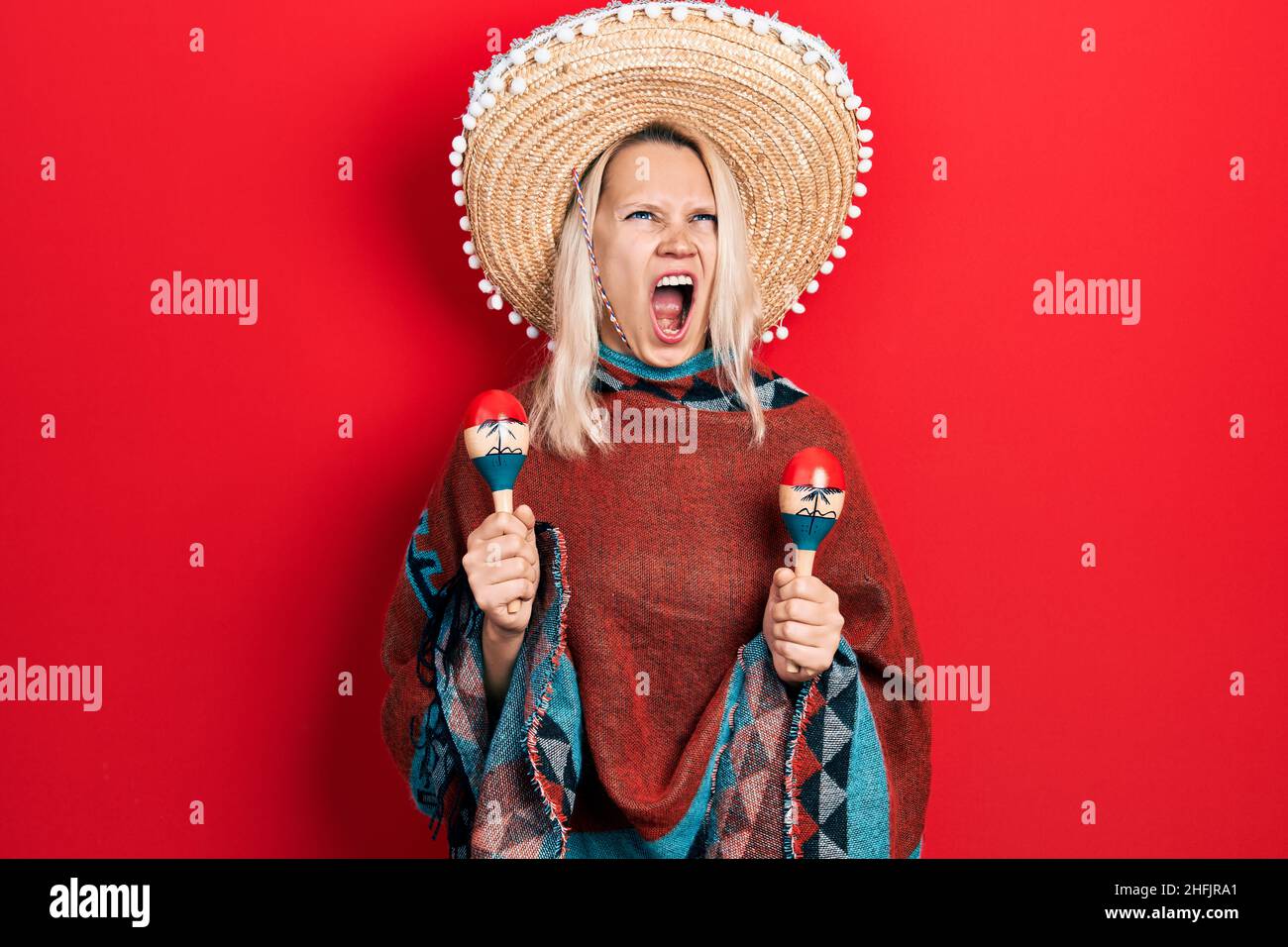 Beautiful caucasian blonde woman wearing festive mexican poncho and maracas angry and mad ...