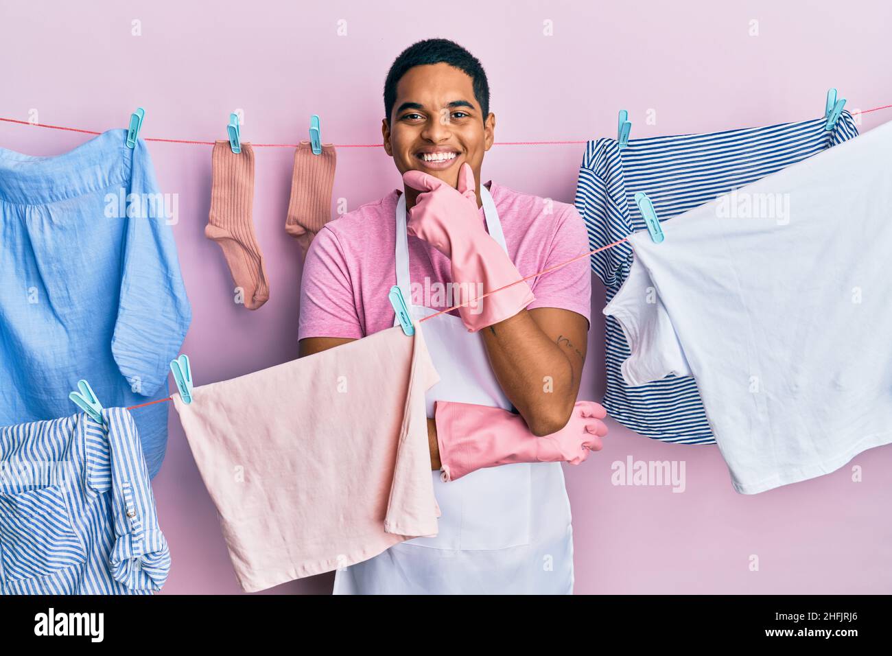 Young handsome hispanic man wearing cleaner apron holding clothes on ...