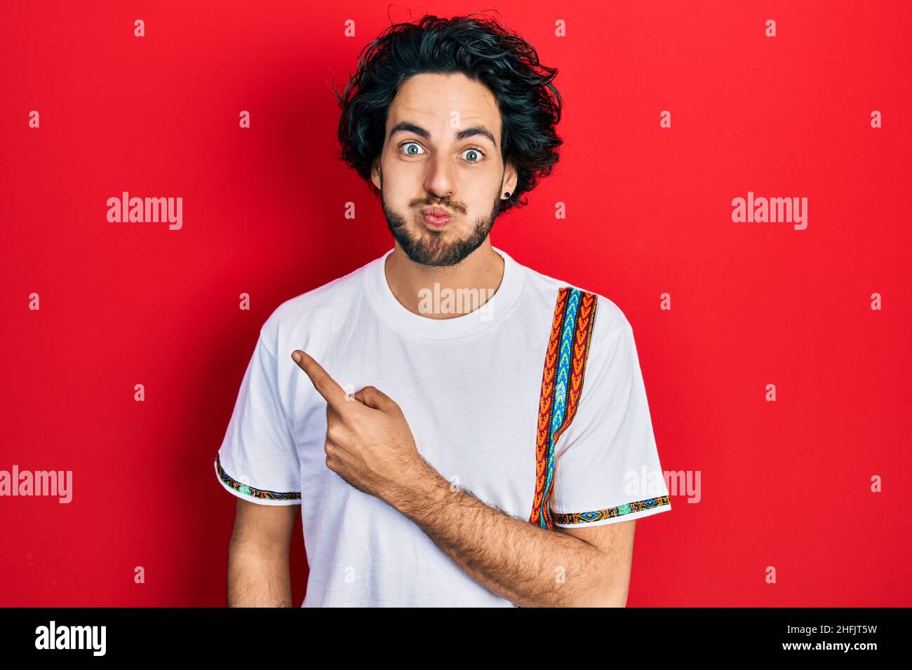 Handsome hispanic man pointing with fingers to the side puffing cheeks ...