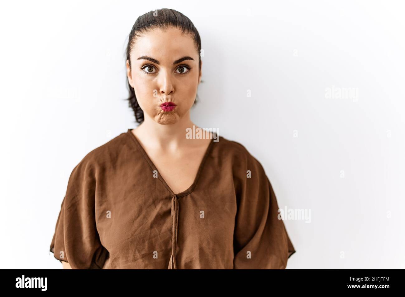 Young brunette woman standing over isolated background puffing cheeks ...