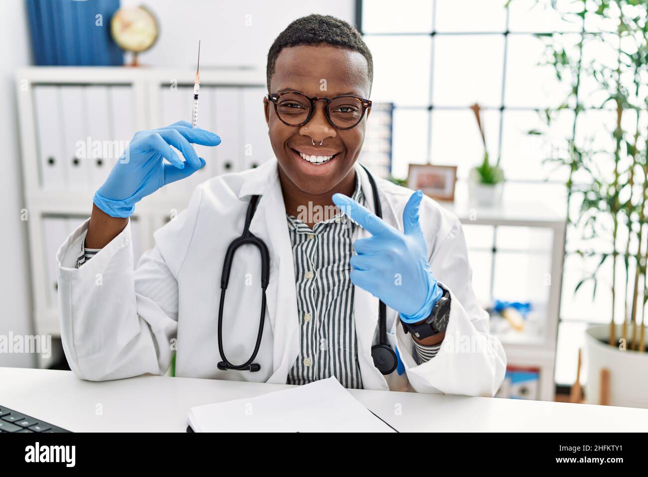 Young african doctor man holding syringe at the hospital cheerful with a smile on face pointing ...