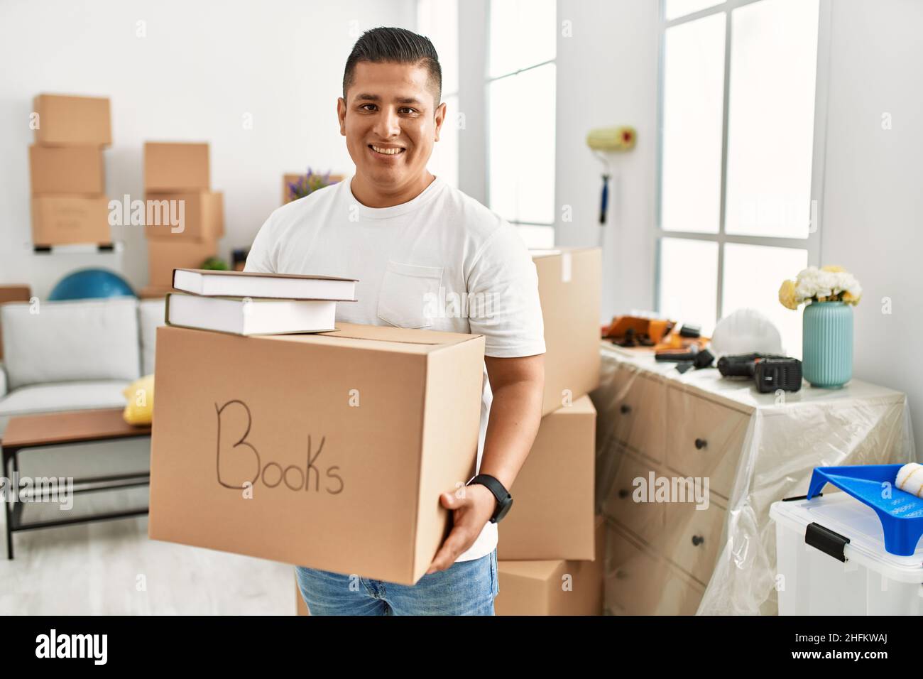 Young latin man smiling happy holding books cardboard box moving at new ...