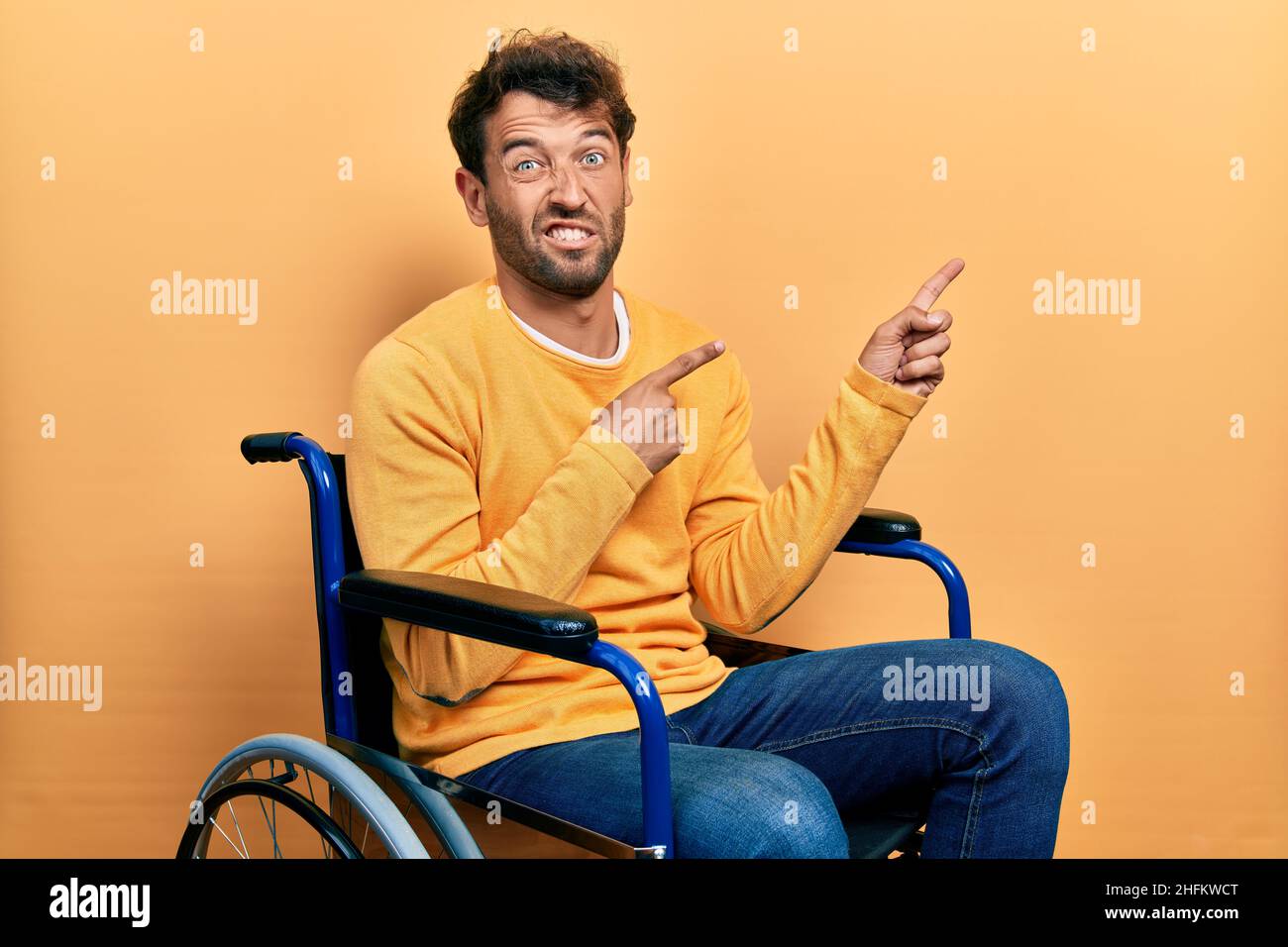 Handsome man with beard sitting on wheelchair pointing aside worried ...