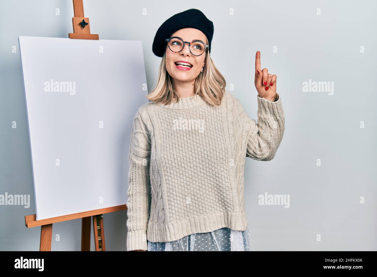 Beautiful caucasian woman standing by painter easel stand pointing ...