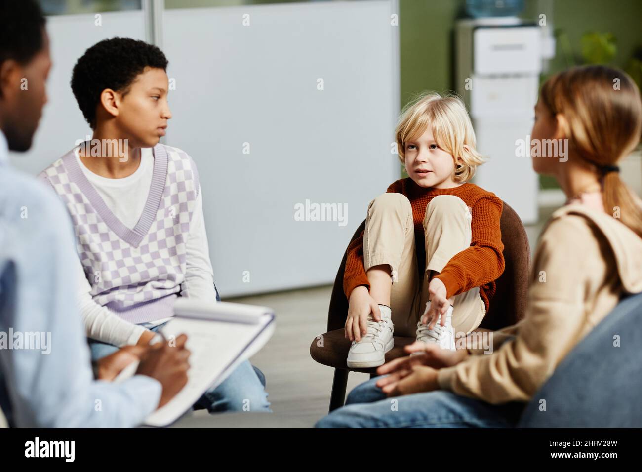 Portrait of cute blond boy speaking in therapy session with diverse ...